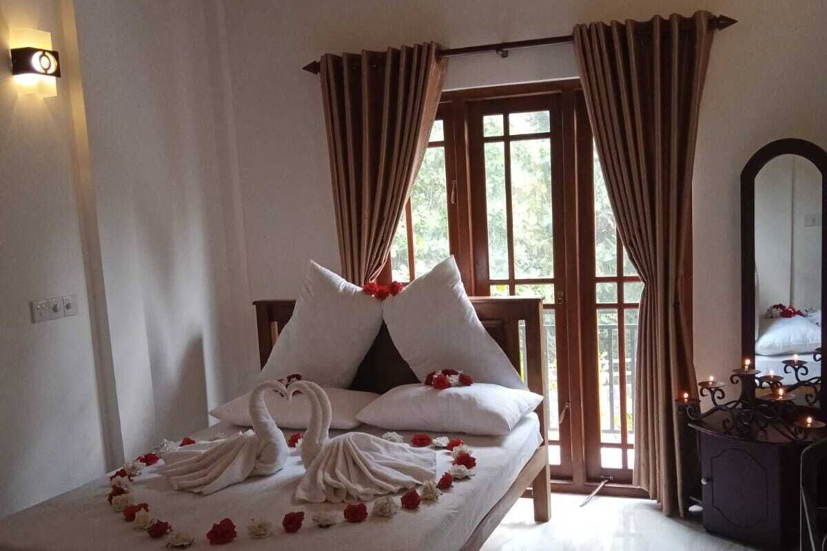 Cama bem arrumada com desenho de cisne e pétalas de rosa, um banco de madeira, um espelho e uma janela com cortinas.