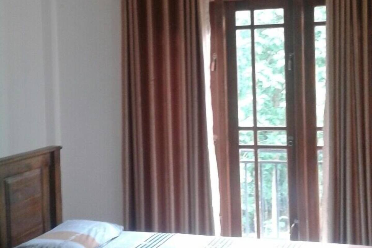 Quarto com cama, ar condicionado, janela com cortinas e vista para o verde lá fora.