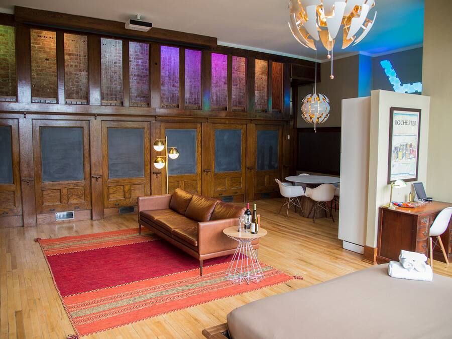 Quarto de hotel com parede revestida de madeira, sofá de couro, mesa redonda com abajur, cama e escrivaninha com computador.