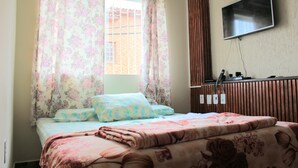 Quarto com cama, cortinas florais, televisão na parede e janela com vista para um edifício de tijolos.