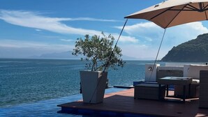 Piscina infinita com mobiliário de exterior e um grande guarda-sol, com vista para um mar calmo e montanhas ao longe.