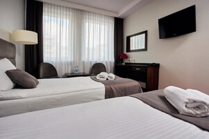 Quarto de hotel com duas camas, uma escrivaninha, uma cadeira, televisão e janela com cortinas.
