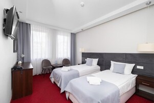 Quarto de hotel com duas camas, uma mesa, uma cadeira, uma televisão e uma janela com cortinas.