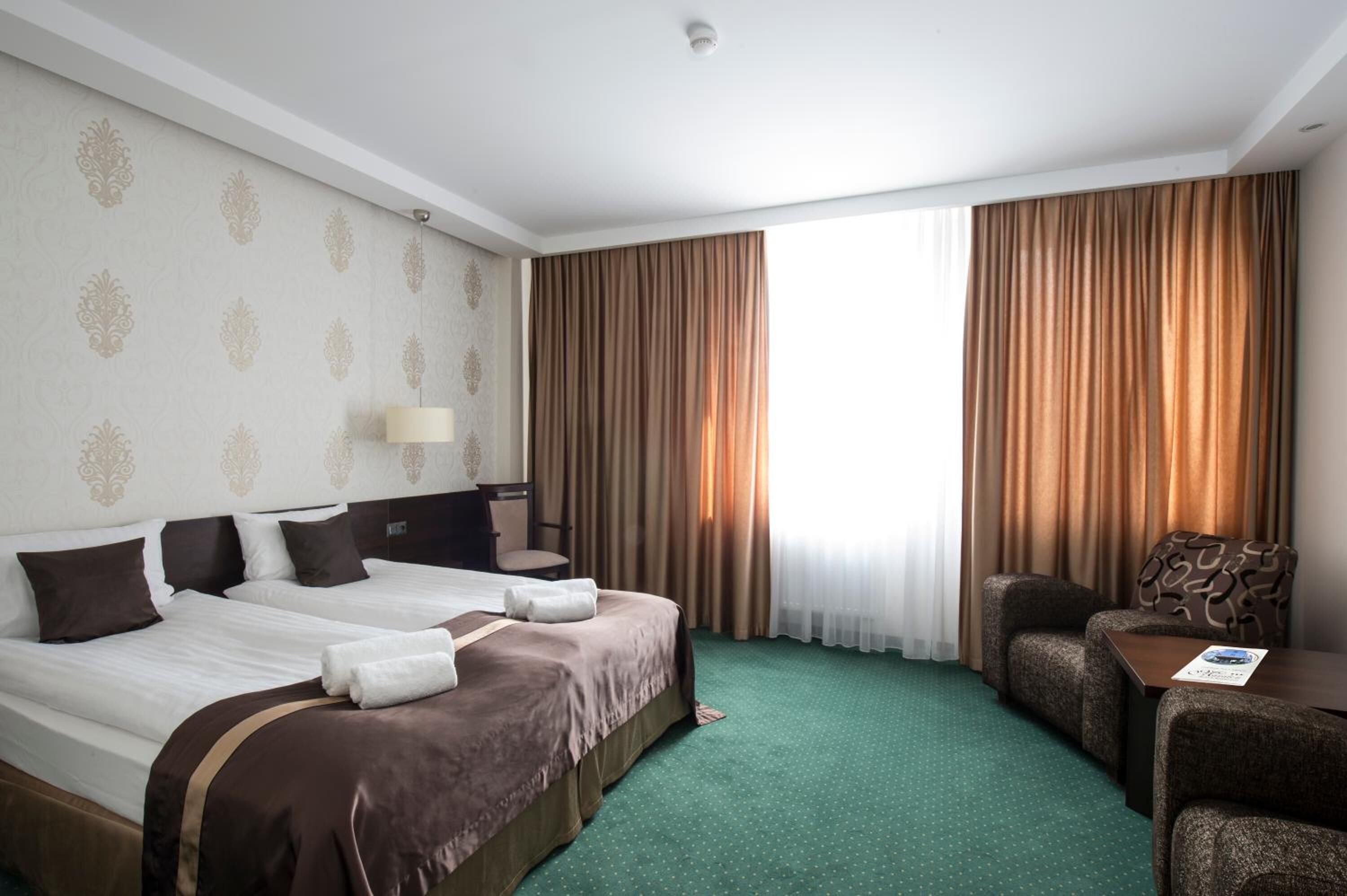 Quarto de hotel com duas camas, uma escrivaninha, uma cadeira e uma janela grande com cortinas.