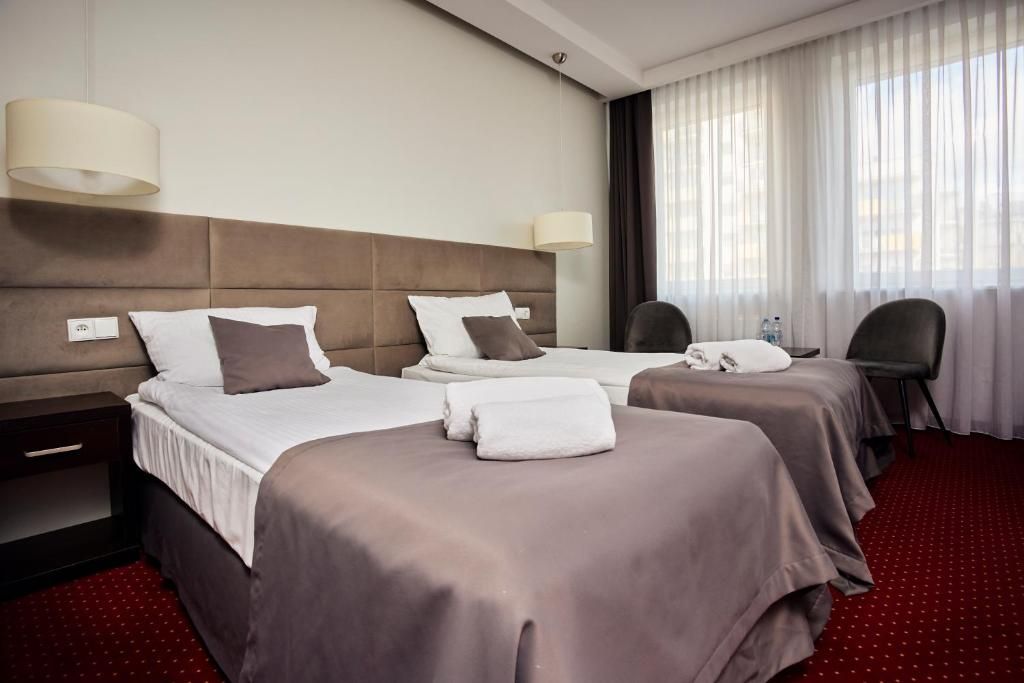 Quarto de hotel com duas camas, um tapete vermelho, uma janela grande com cortinas e uma luminária na parede.