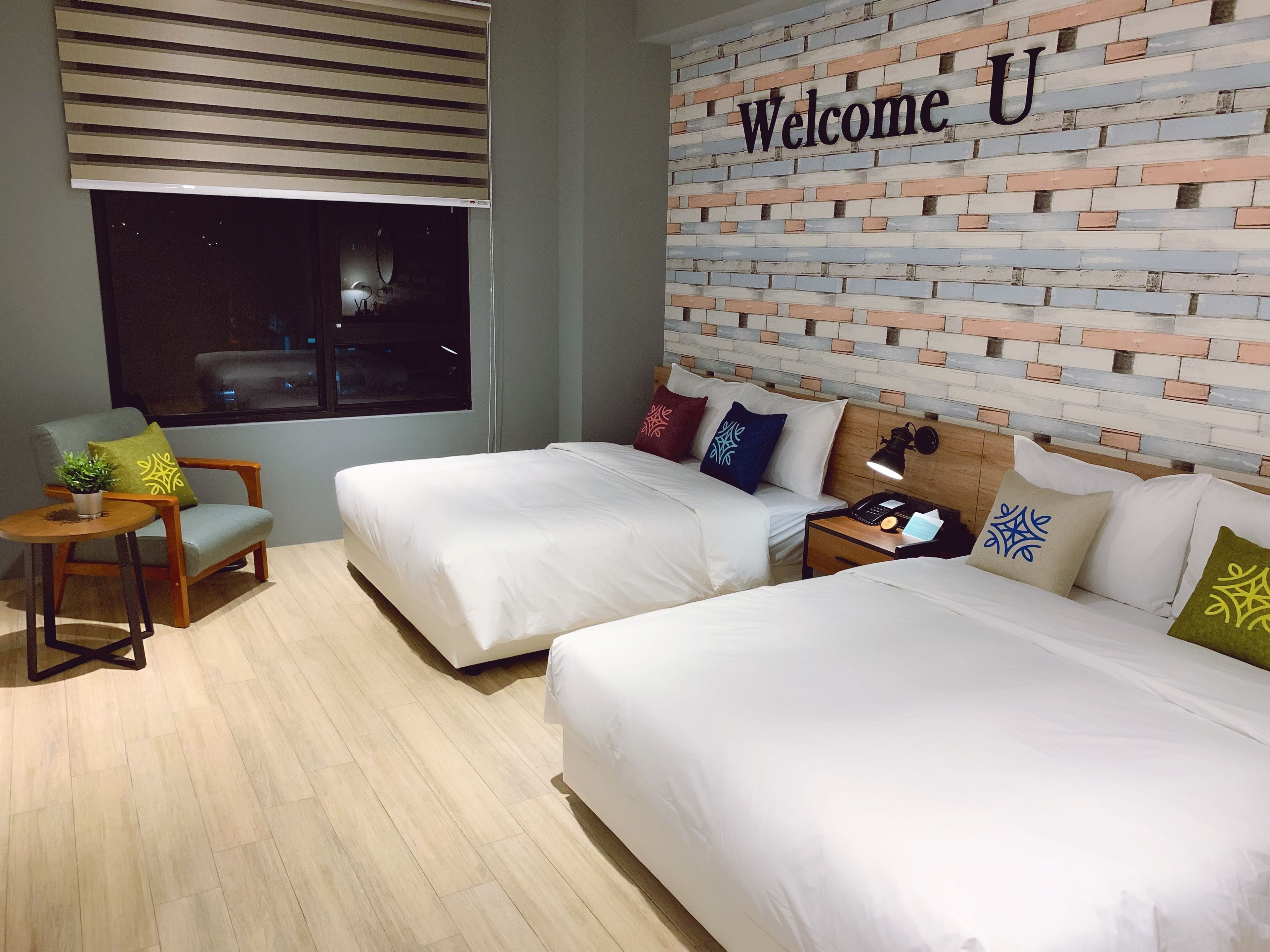 Quarto de hotel com duas camas, uma mesinha com uma planta, uma cadeira e uma parede com a placa 'Welcome'.