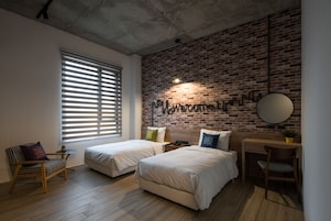 Quarto moderno com duas camas, uma mesa e uma cadeira. Uma parede de tijolos com um letreiro de 'Welcome'.