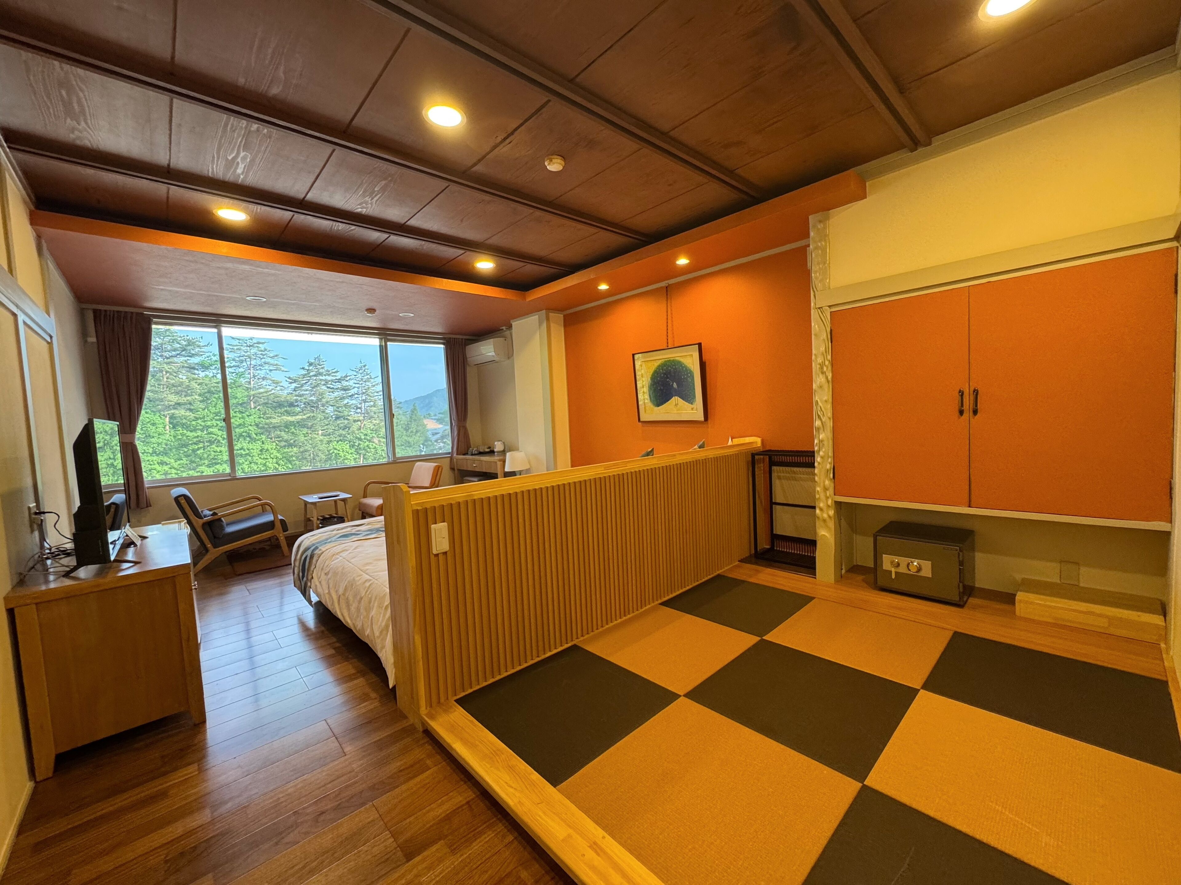 Quarto de hotel com cama, escrivaninha, cadeira, janela com vista para árvores e parede laranja.