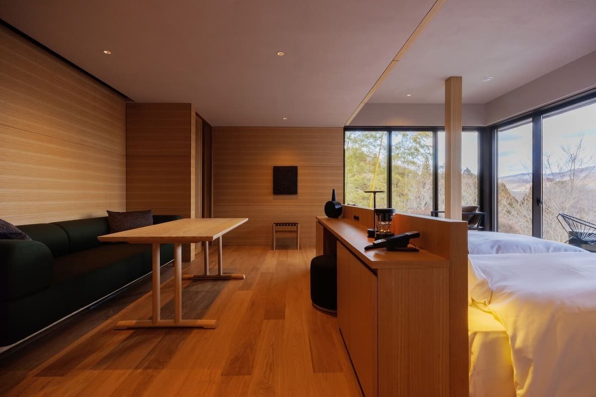 Quarto moderno com piso de madeira, sofá verde, escrivaninha de madeira, cama e grandes janelas com vista para árvores.