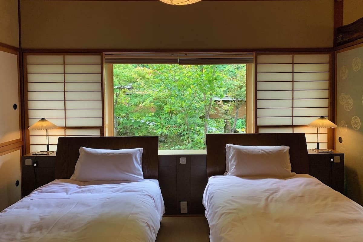 Quarto tradicional japonês com duas camas, biombos shoji, piso de esteiras de tatame e vista para o verde lá fora.