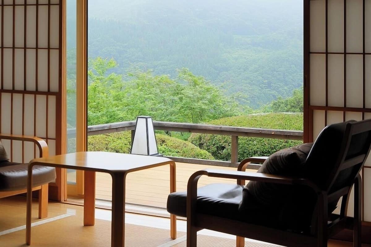 Quarto com piso de tatame, uma mesa baixa, uma cadeira e vista para uma cadeia montanhosa arborizada.