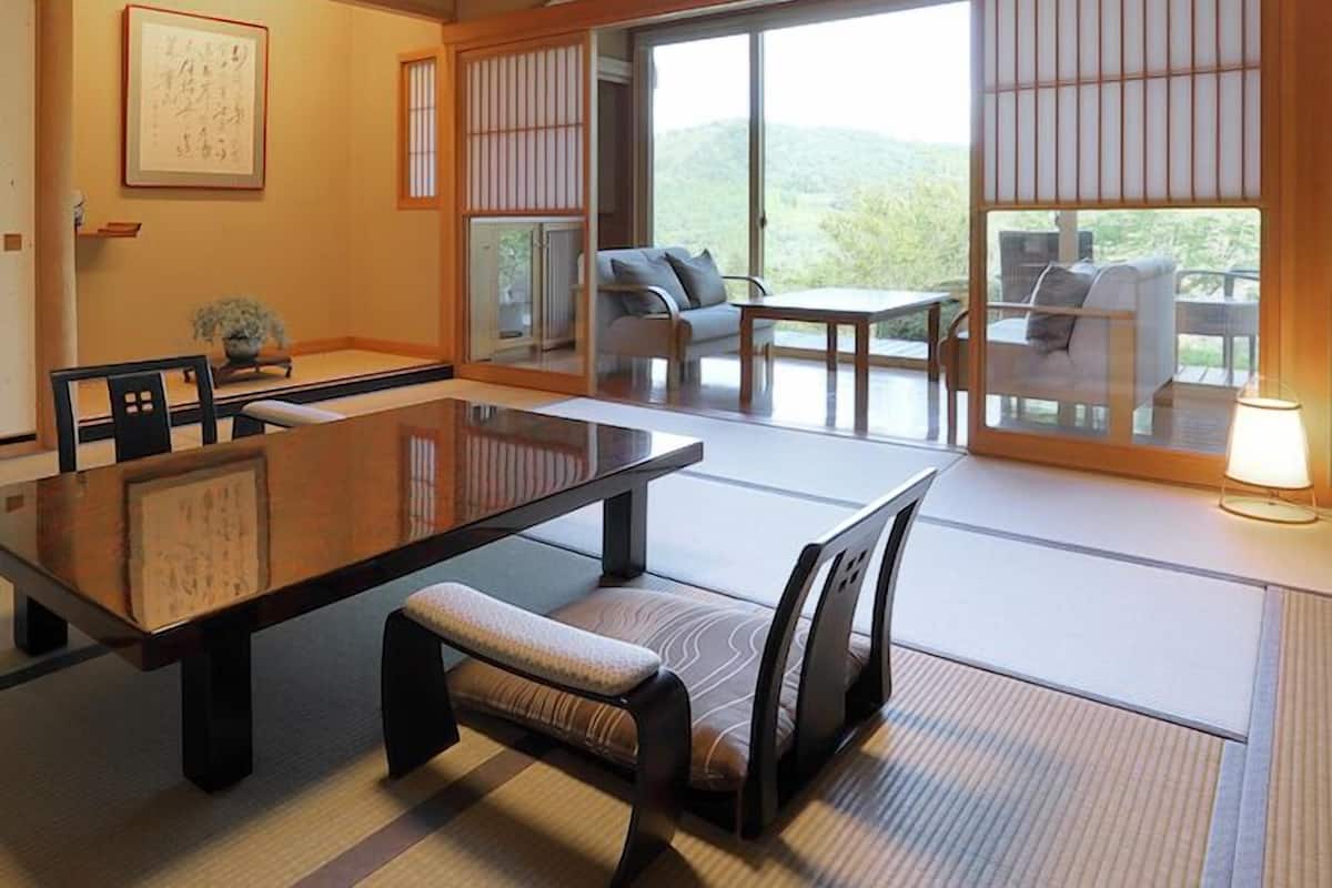 Quarto tradicional em estilo japonês, com piso de tatame, uma mesa baixa e portas deslizantes que dão para uma varanda com vista para as colinas.