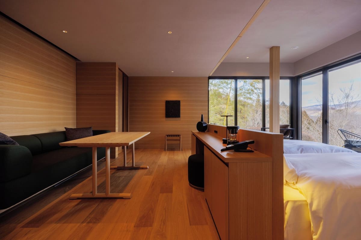 Quarto moderno com piso de madeira, sofá verde, escrivaninha de madeira, cama e janelas amplas.