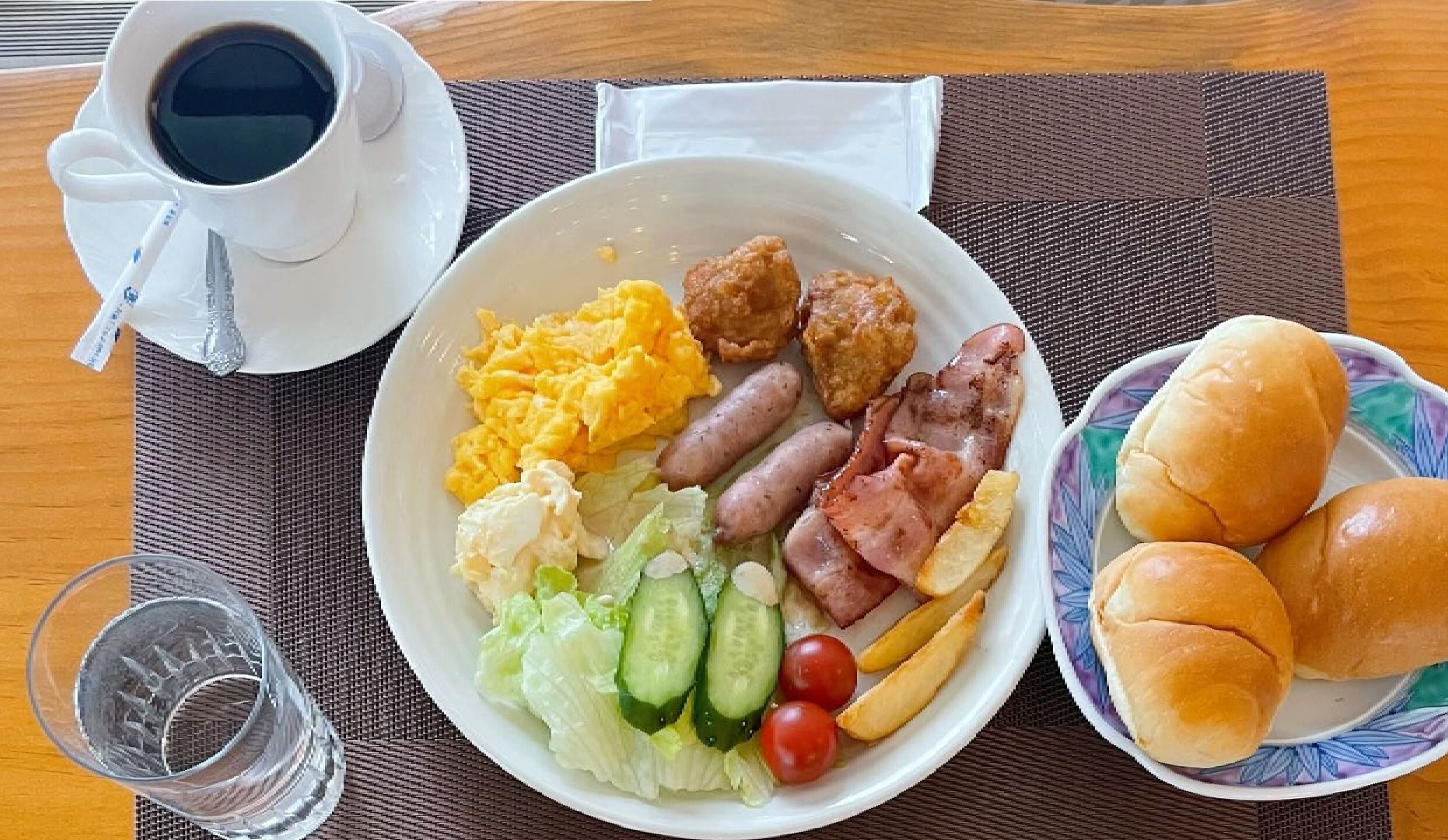 Prato de café da manhã com ovos mexidos, salsichas, bacon e uma salada acompanhado de uma xícara de café e um copo de água sobre uma mesa.