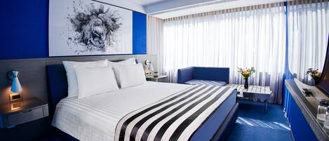 Quarto de hotel moderno com uma cama grande, uma mesinha de cabeceira com abajur, um pequeno espaço para sentar e uma janela ampla com cortinas.