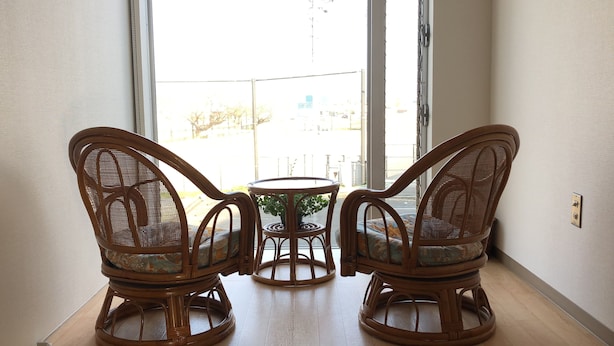 Quarto com duas cadeiras de vime, uma pequena mesa, uma janela com vista para a rua e um interruptor na parede.