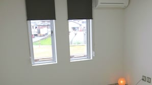 Quarto com tatames, duas janelas com persianas escuras, um aparelho de ar condicionado e uma luminária fixada na parede.