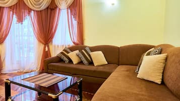 Uma sala de estar com sofá marrom, almofadas estampadas, mesa de centro de vidro e cortinas leves.