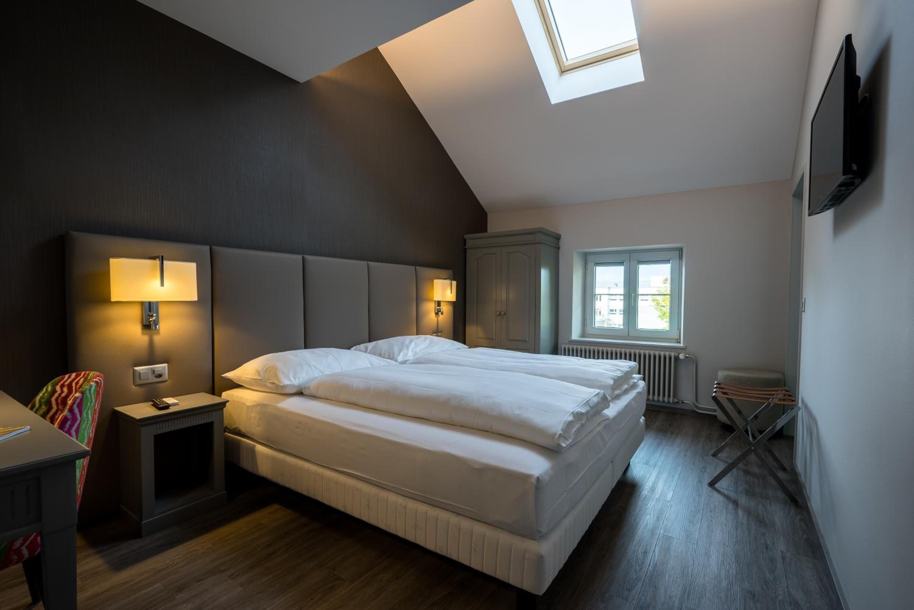 Quarto moderno com claraboia, cama, abajures, radiador e televisão na parede.