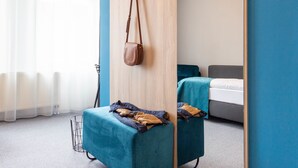 Quarto de hotel com um espelho grande, um guarda-roupa de madeira, uma cadeira azul e uma pequena mesa com uma bolsa pendurada no guarda-roupa.