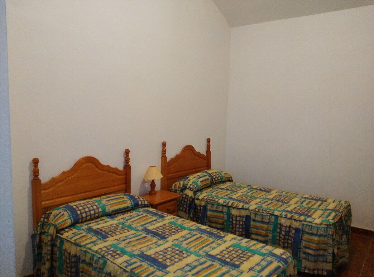 Quarto com duas camas de madeira, cada uma com colcha estampada, uma mesinha de cabeceira e abajur, e parede branca lisa.