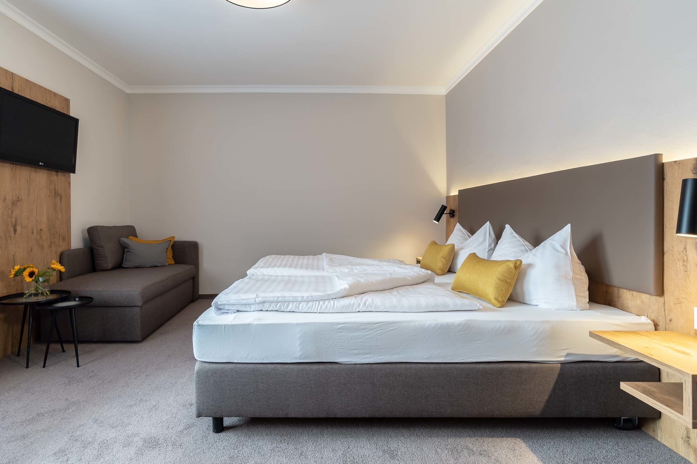 Quarto moderno com cama, sofá, mesinha com flores e TV na parede.