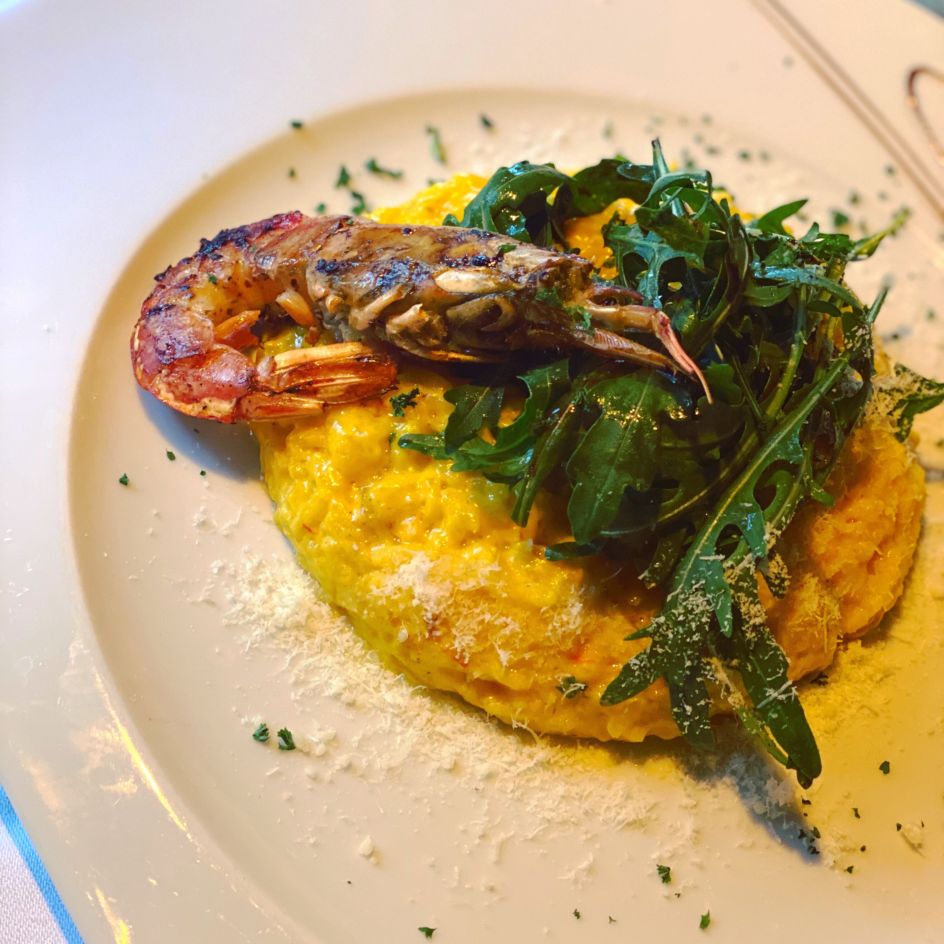 Um prato com um prato de frutos do mar, possivelmente lagosta, servido sobre um leito de risoto amarelo, guarnecido com vegetais e queijo ralado.