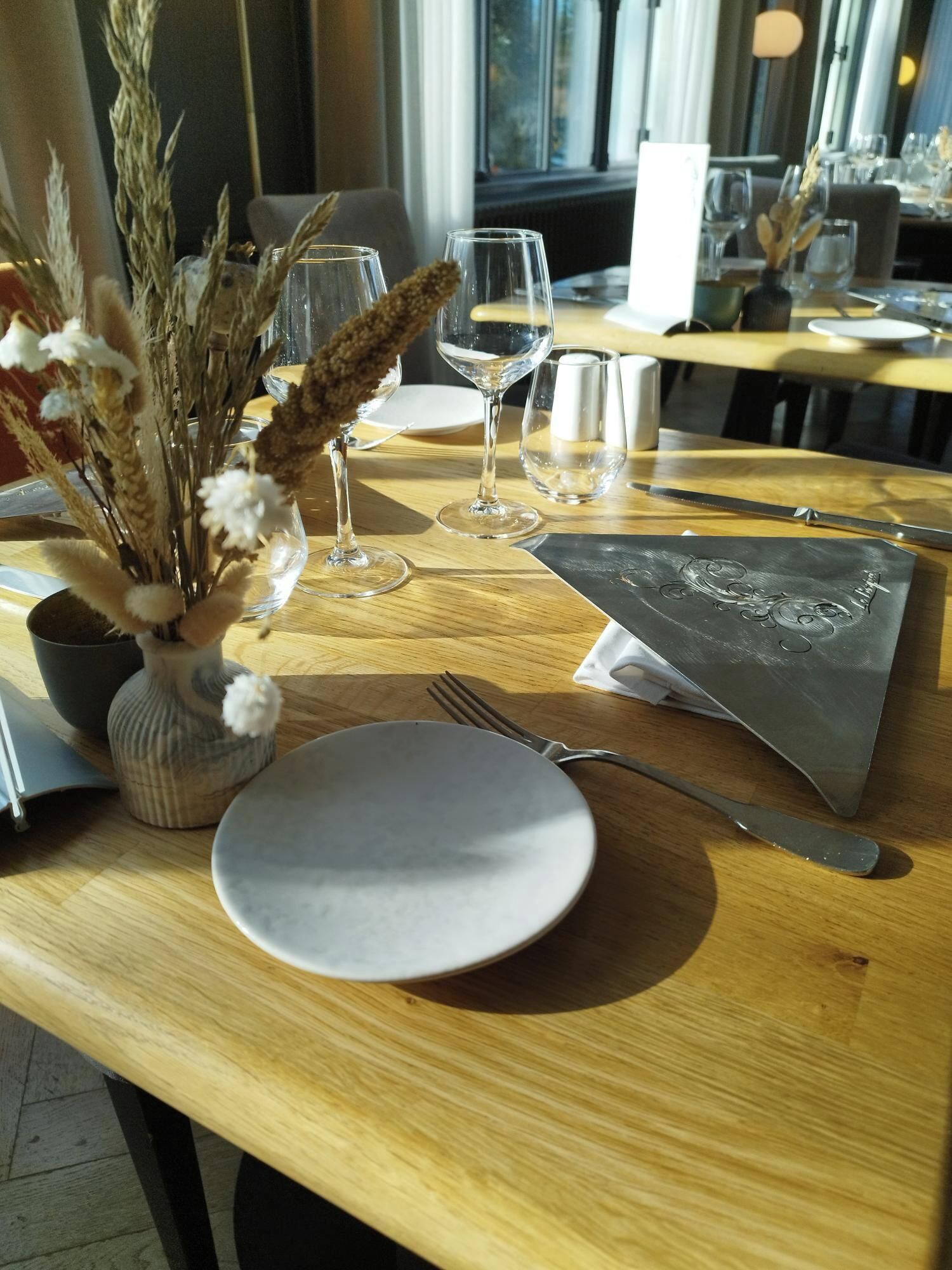 Uma mesa de jantar posta com prato, garfo, faca e um vaso decorativo com flores secas.