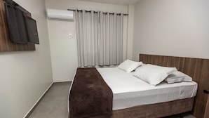 Cama bem arrumada com lençóis brancos e um cobertor castanho, ar condicionado fixado na parede e uma cortina.