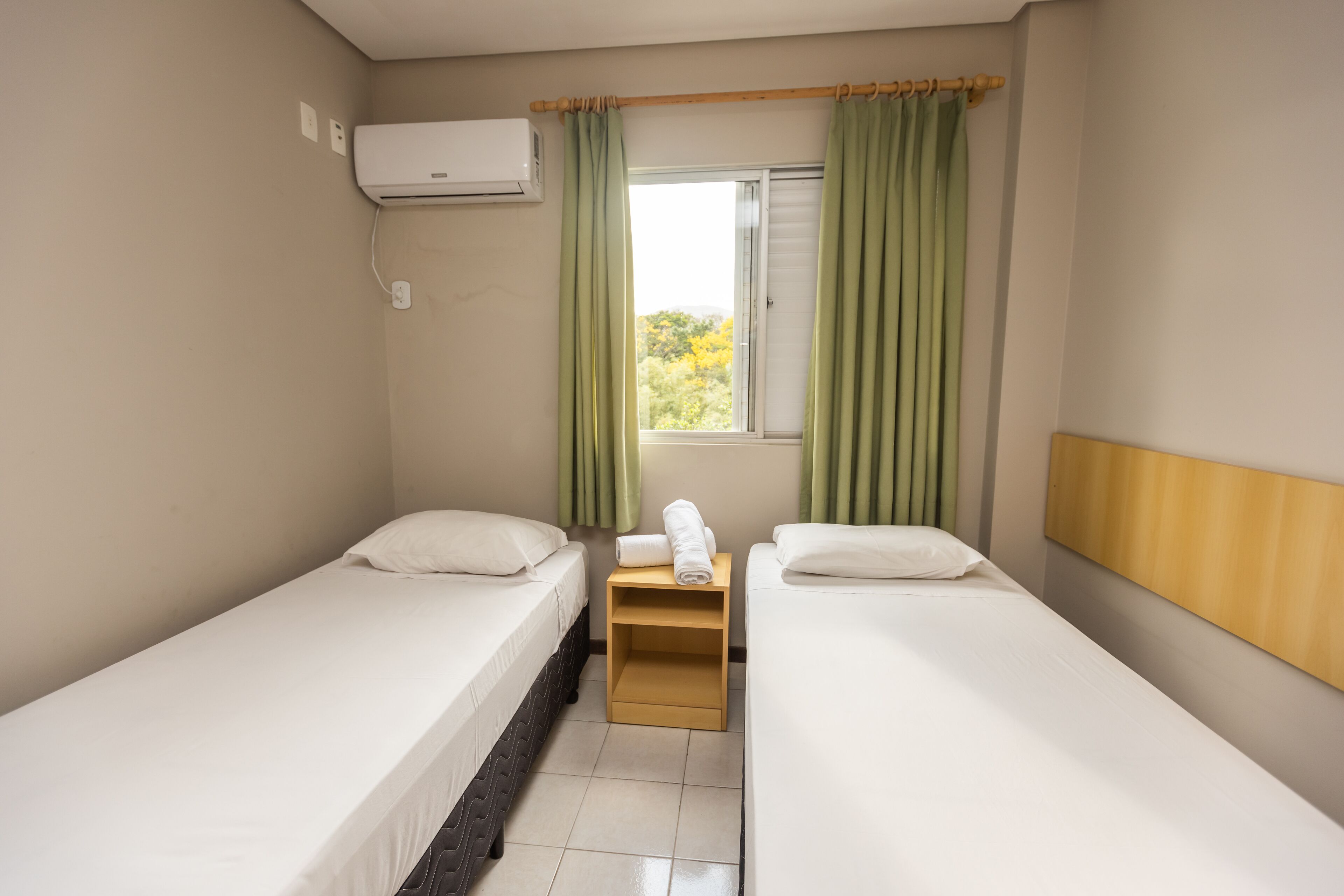Quarto de hotel com duas camas de solteiro, uma mesinha de cabeceira de madeira, uma janela com cortinas, um aparelho de ar condicionado e vista para árvores.