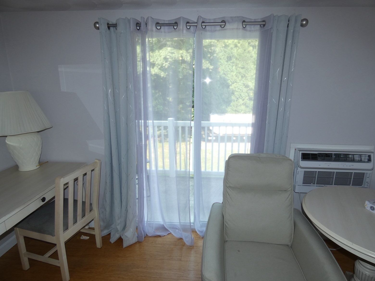 Quarto com uma mesa branca, uma cadeira, um abajur, uma janela com cortinas e um aparelho de ar condicionado.