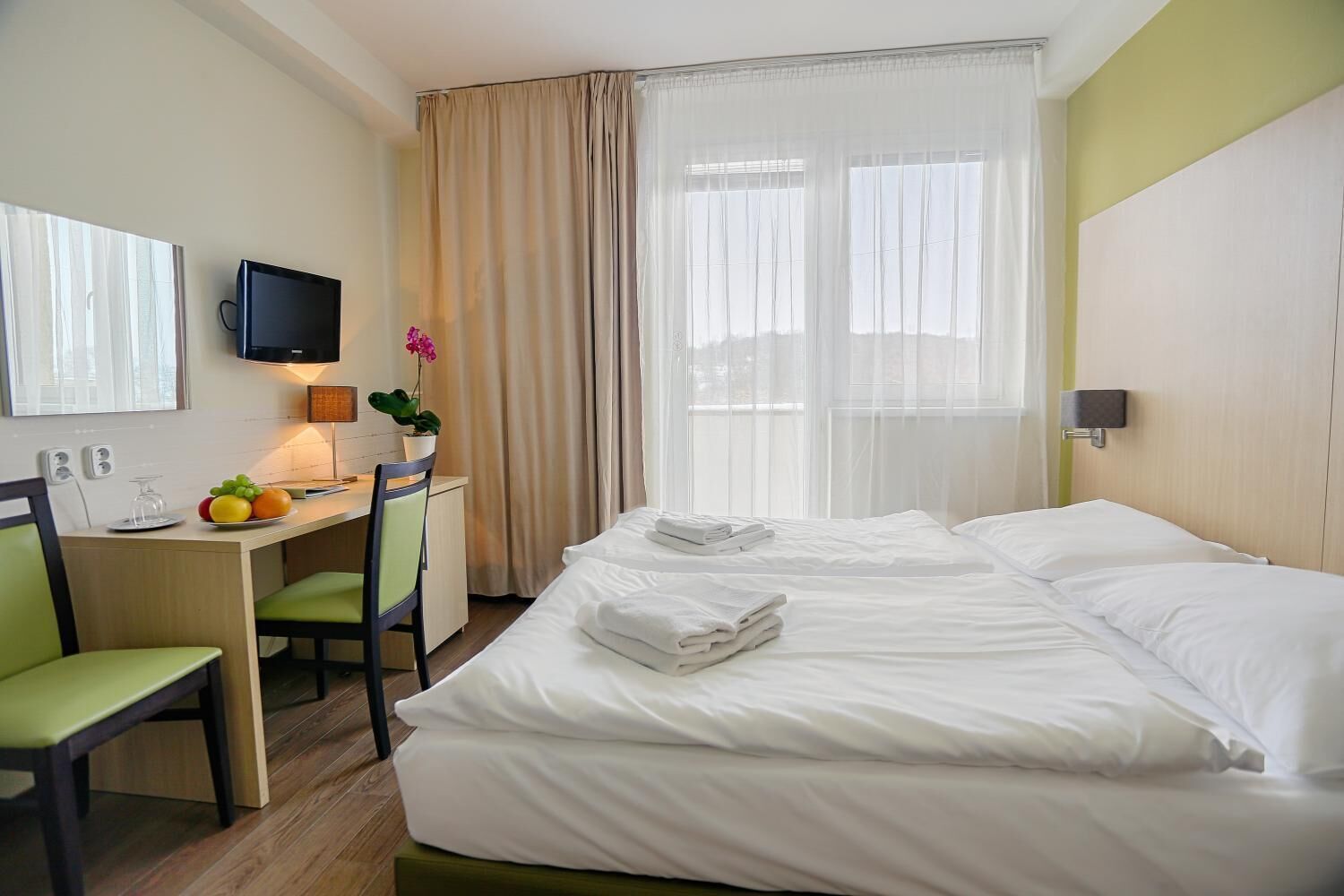 Quarto de hotel com cama, escrivaninha com cadeira, televisão e janela com cortinas.