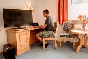 Um homem trabalha numa secretária de madeira com um laptop, uma televisão e um altifalante. Há uma cadeira com padrão floral e uma mesa pequena com um vaso de flores.
