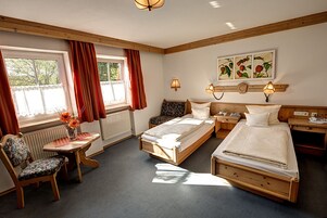Quarto de hotel com duas camas, uma mesa, uma cadeira, um radiador e uma janela com cortinas.