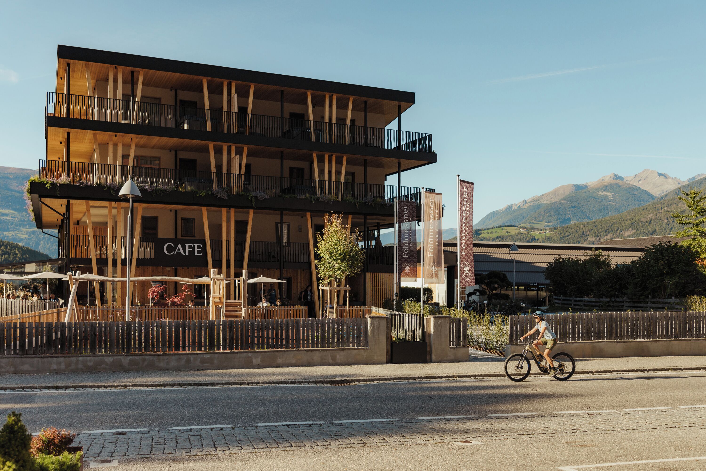 Edifício de vários andares com café no andar térreo, mesas ao ar livre e vista para as montanhas.