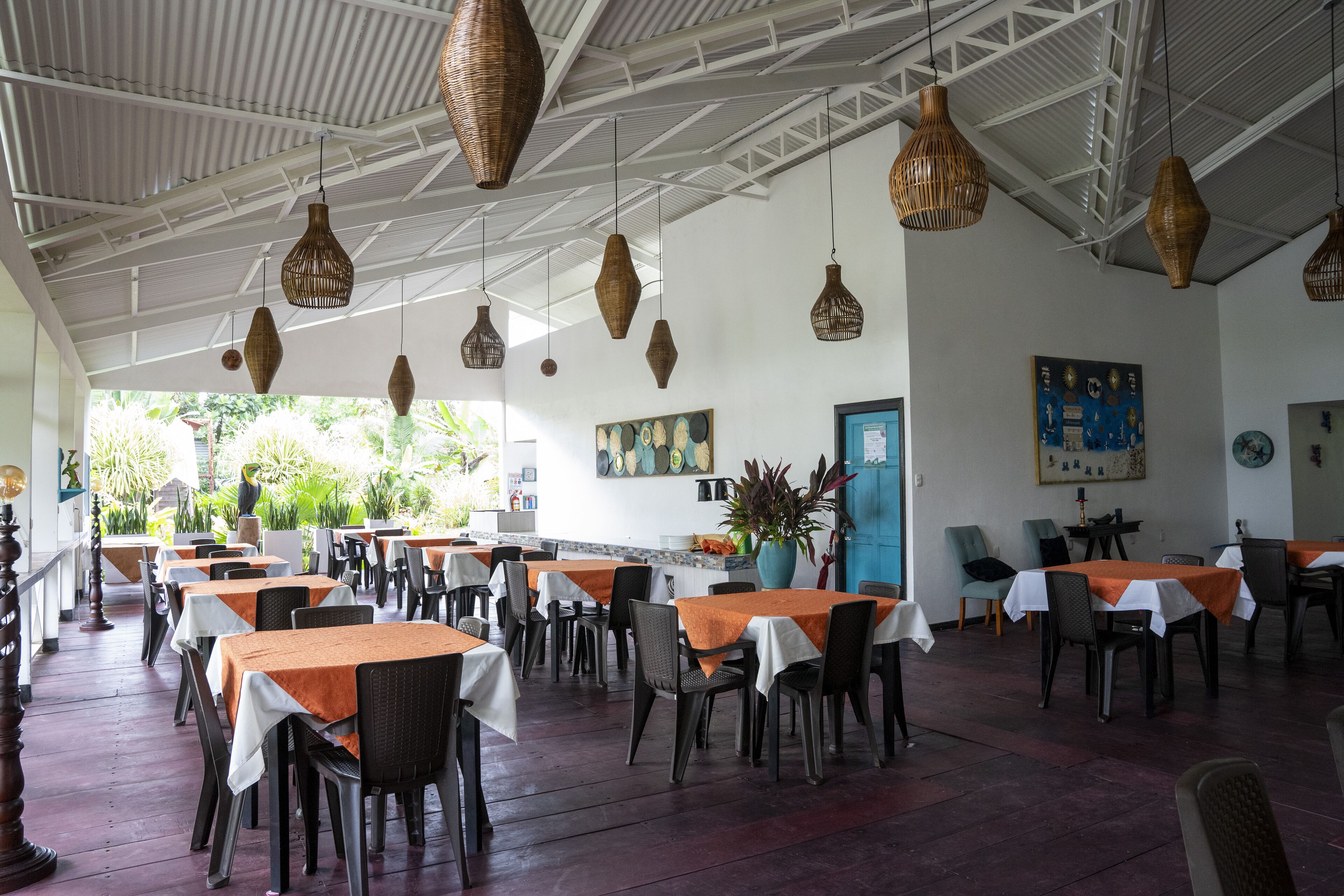 Restaurante com várias mesas cobertas por toalhas laranja e branco, luminárias de vime suspensas e vista para o verde lá fora.