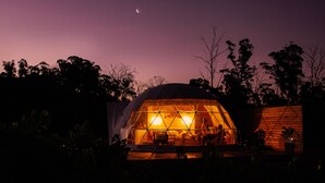 Uma estrutura em forma de cúpula com fachada de vidro, iluminação interna e vista do céu noturno com uma lua crescente.