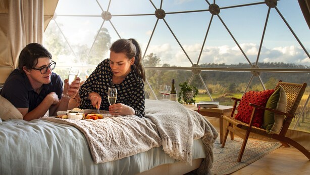Um casal jantando em uma sala em formato de cúpula geodésica, com vista para o campo.