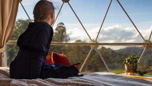 Uma pessoa sentada em uma cama, com vista para uma paisagem montanhosa através de uma estrutura de janela geométrica.