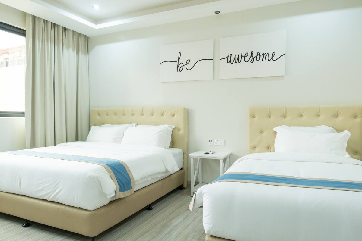 Quarto de hotel com duas camas, uma pequena mesa de cabeceira e uma parede decorada com mensagens motivacionais.
