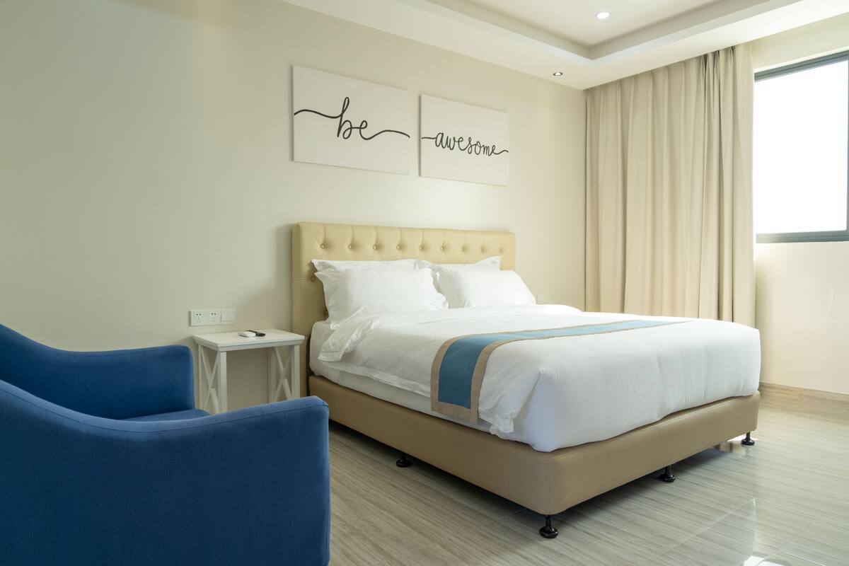 Quarto de hotel com uma cama grande, uma poltrona azul, uma pequena mesa de cabeceira e duas obras de arte nas paredes.