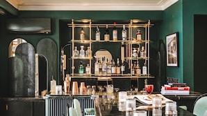 Uma área de bar com um balcão comprido, várias prateleiras cheias de garrafas e um espaço de estar com cadeiras e uma mesa.