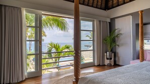 Quarto com uma grande janela, cama de madeira, planta em vaso e varanda com vista para o mar.