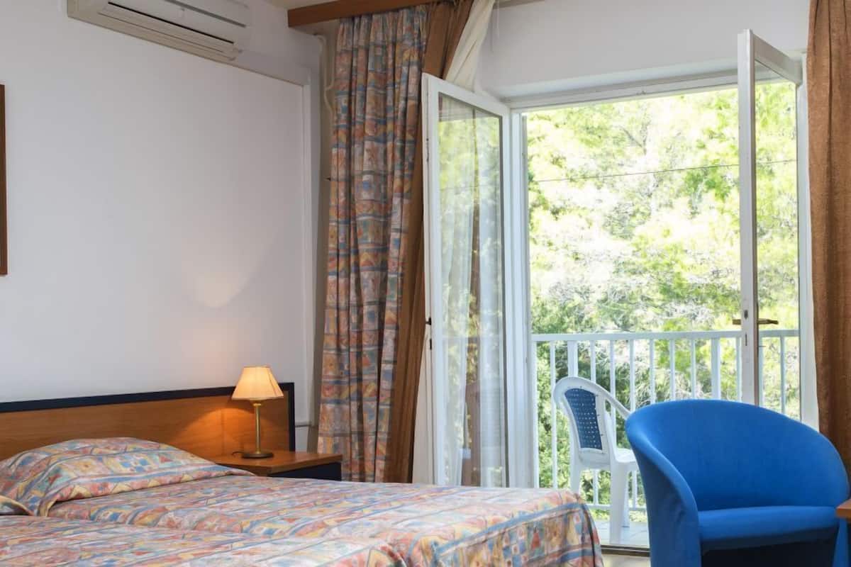 Quarto de hotel com cama, cadeira, luminária e varanda com vista para árvores.