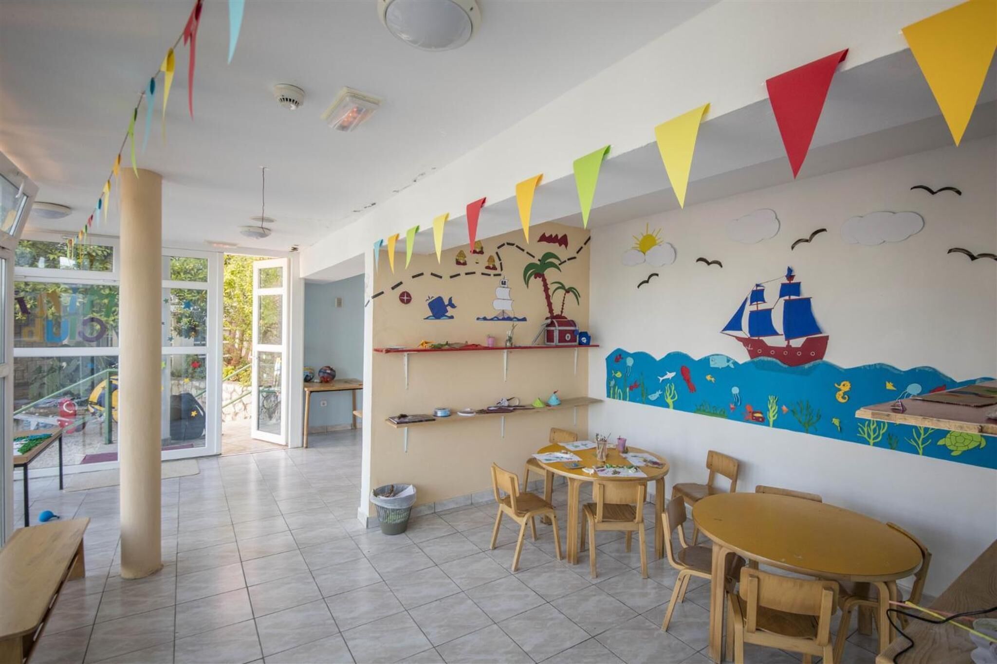 Espaço infantil com mural, mesas, cadeiras e um contentor de lixo.