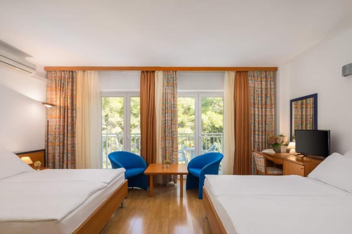 Quarto de hotel com duas camas, uma mesa, uma cadeira, uma televisão e janelas amplas com cortinas.