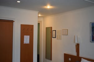 Quarto de hotel com porta de madeira, espelho, cama com cabeceira e luminária fixada na parede.