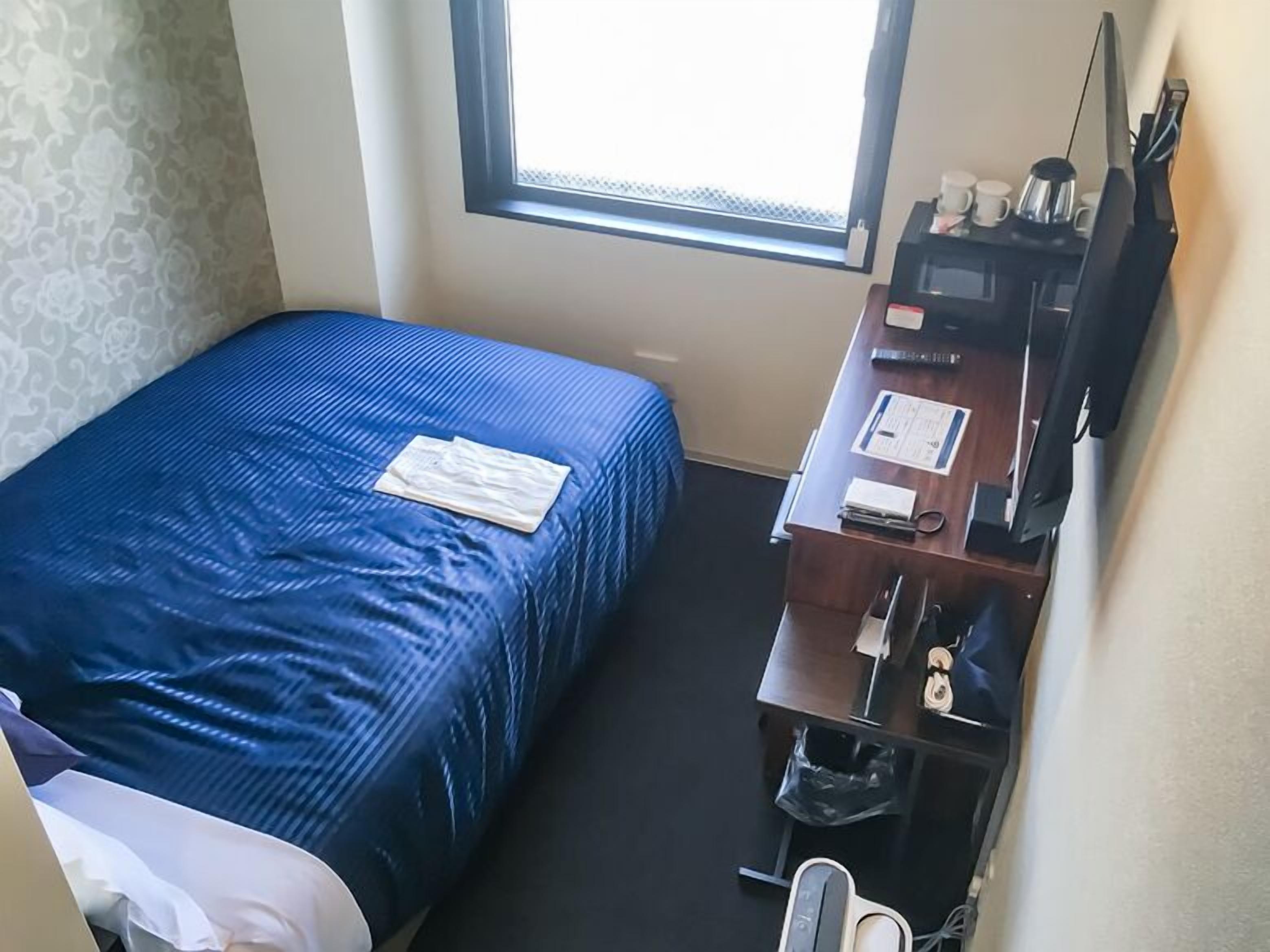 Quarto de hotel com cama, escrivaninha com computador, cadeira e janela.