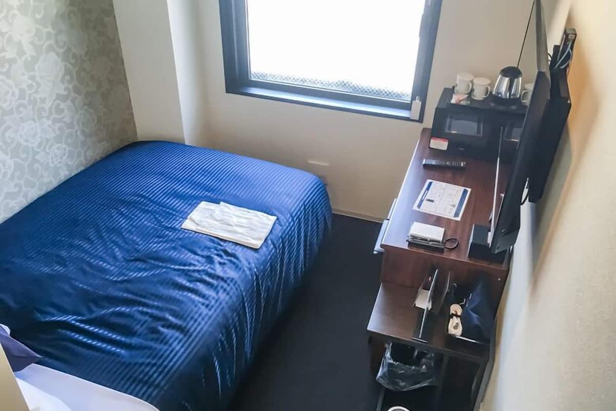 Quarto de hotel com cama, escrivaninha com computador, cadeira e janela.