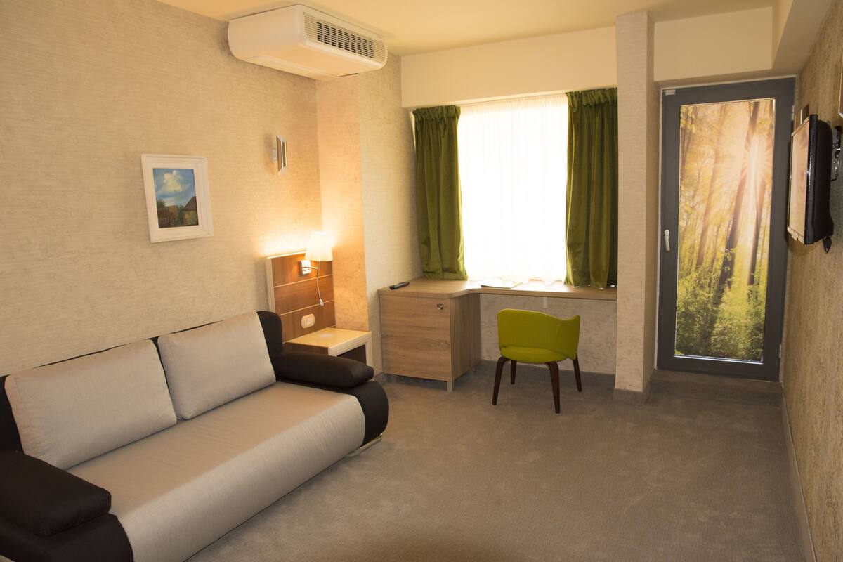 Quarto de hotel com sofá, escrivaninha, cadeira, quadro na parede e janela com cortinas.